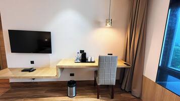 Quarto de hotel moderno com televisão de tela plana, escrivaninha de madeira, cadeira estofada, luminária pendente e janela com cortinas.