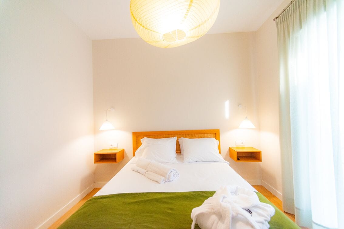Quarto de hotel com uma cama grande, duas mesas de cabeceira, um candeeiro pendente e uma janela com cortinas.