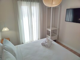 Cama bem arrumada com lençóis brancos e toalhas dobradas, mesa de cabeceira branca, abajur, televisão na parede e janela com cortinas translúcidas.
