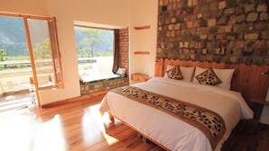 Quarto com cama grande, piso de madeira, parede de pedra e vista através de uma porta deslizante.