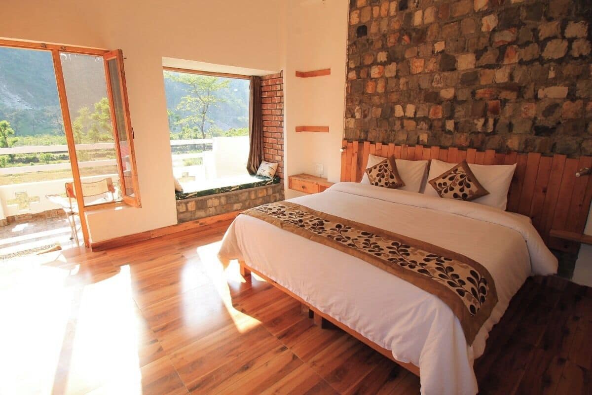 Quarto com cama grande, piso de madeira, parede de pedra e vista através de uma porta deslizante.
