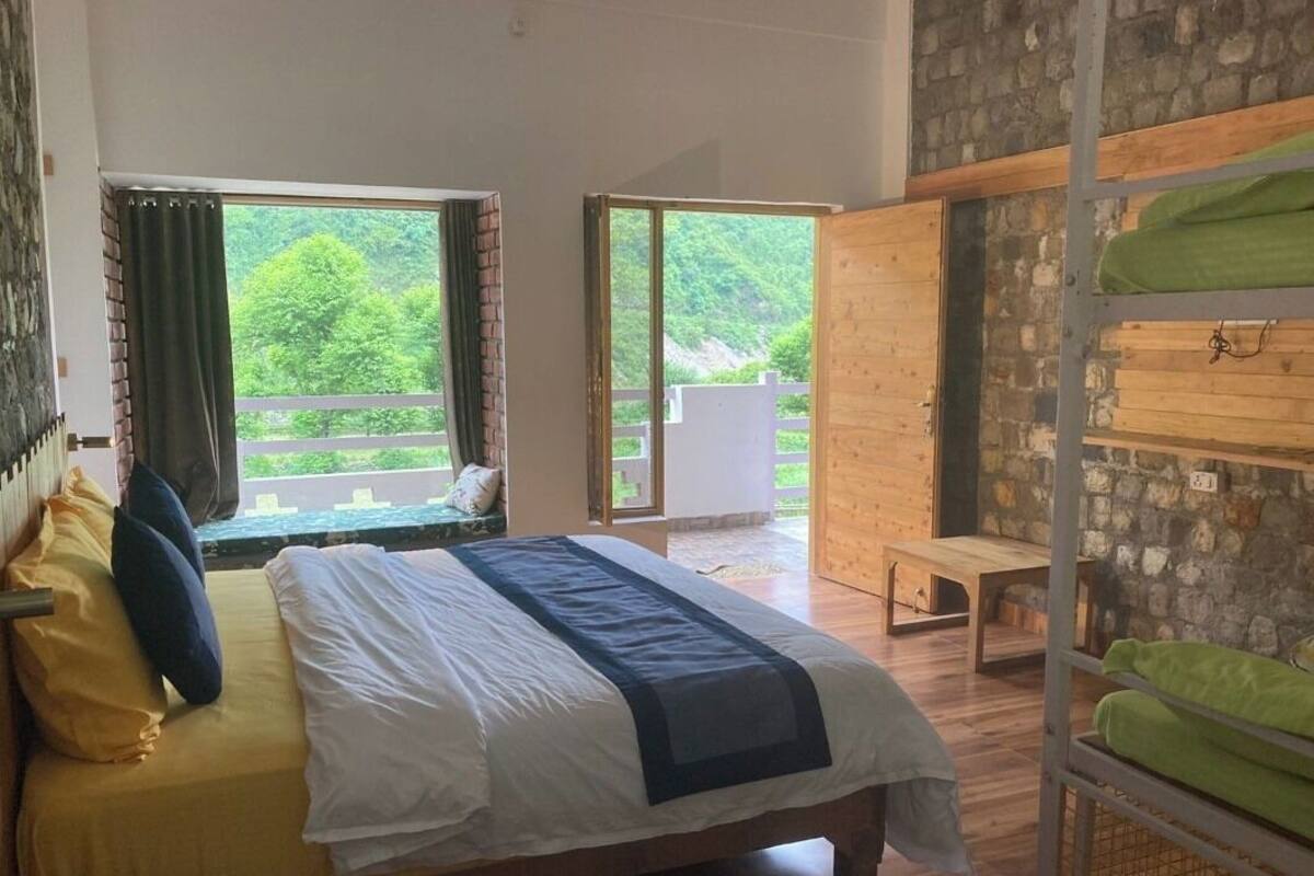 Quarto com cama, um banco, uma beliche e vista para o verde lá fora.