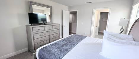 Um quarto com uma cama, uma cômoda, uma televisão sobre a cômoda e uma porta que dá para outro espaço.