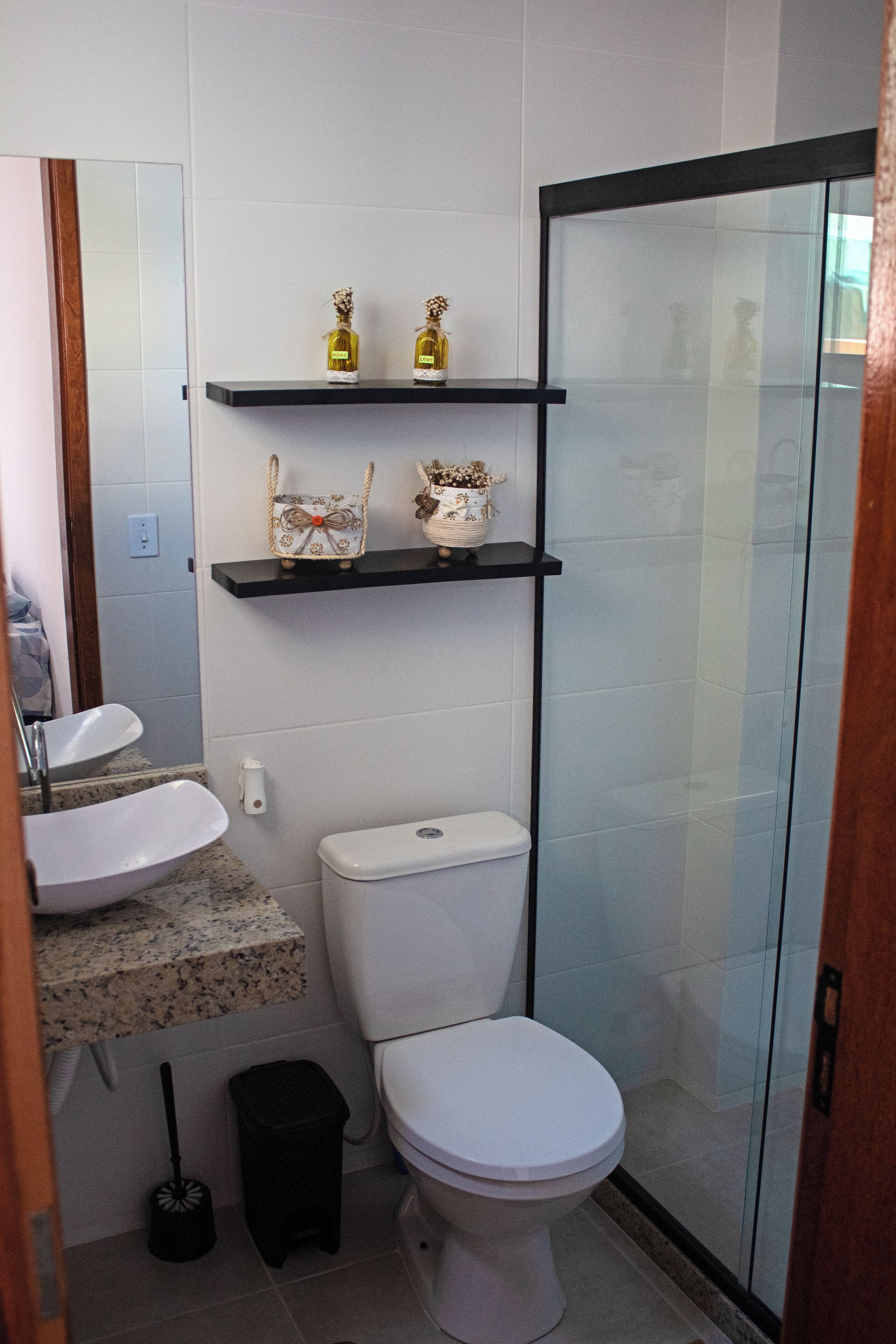 Casa de banho com sanita branca, lavatório, chuveiro e uma prateleira com objetos decorativos.