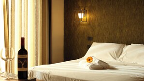 Quarto de hotel com uma cama bem arrumada, uma garrafa de vinho e um copo sobre uma mesa de madeira.