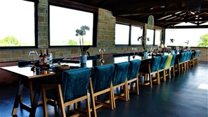 Uma longa mesa de jantar com cadeiras, numa sala com janelas amplas e uma parede de pedra.