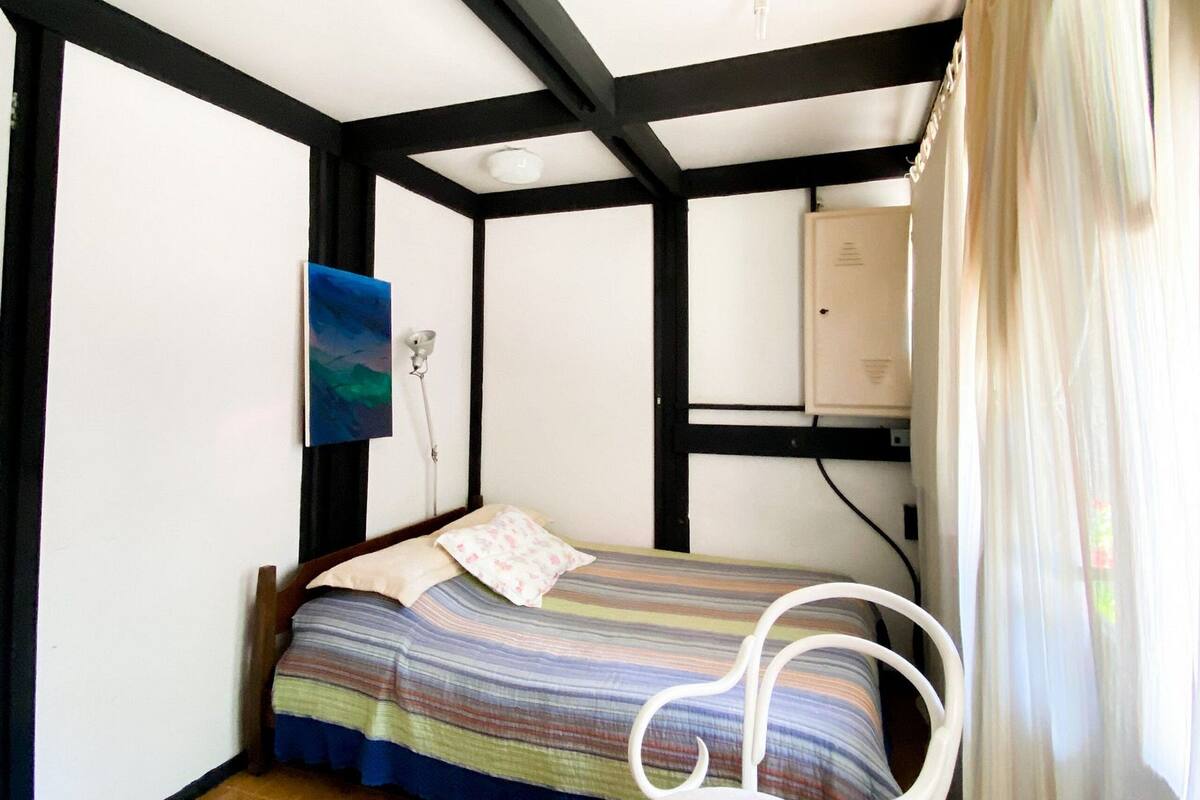 Um quarto pequeno e acolhedor com uma cama de casal, uma cadeira branca e um quadro na parede.