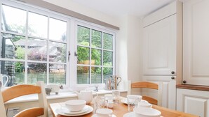 Uma sala de jantar com mesa de madeira posta para quatro pessoas, cadeiras brancas e janela com vista para o verde lá fora.