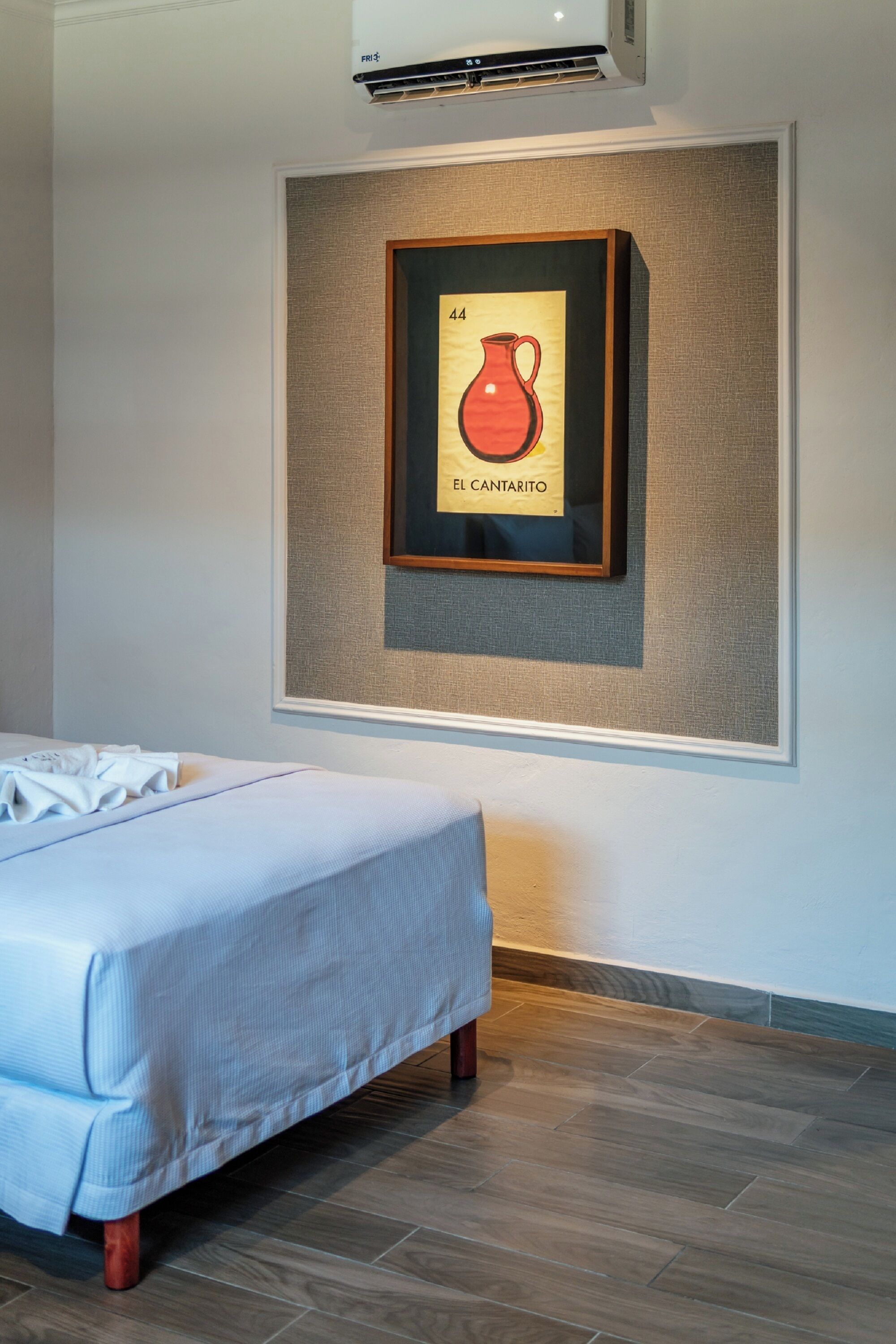 Quarto de hotel com uma cama, uma obra de arte emoldurada de um jarro vermelho com a etiqueta "El Cantario" na parede, e piso de madeira.