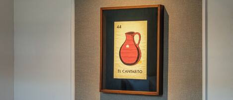 Quarto de hotel com uma cama, uma obra de arte emoldurada de um jarro vermelho com a etiqueta "El Cantario" na parede, e piso de madeira.
