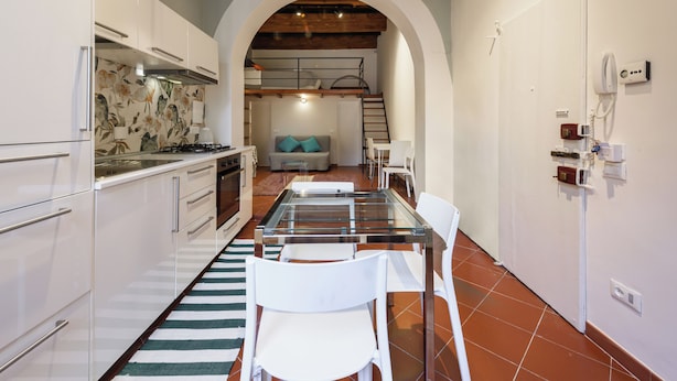 Cozinha moderna com armários brancos, mesa de jantar de vidro e cadeiras brancas. Há uma escada que leva a um quarto no mezanino com cama e sofá.