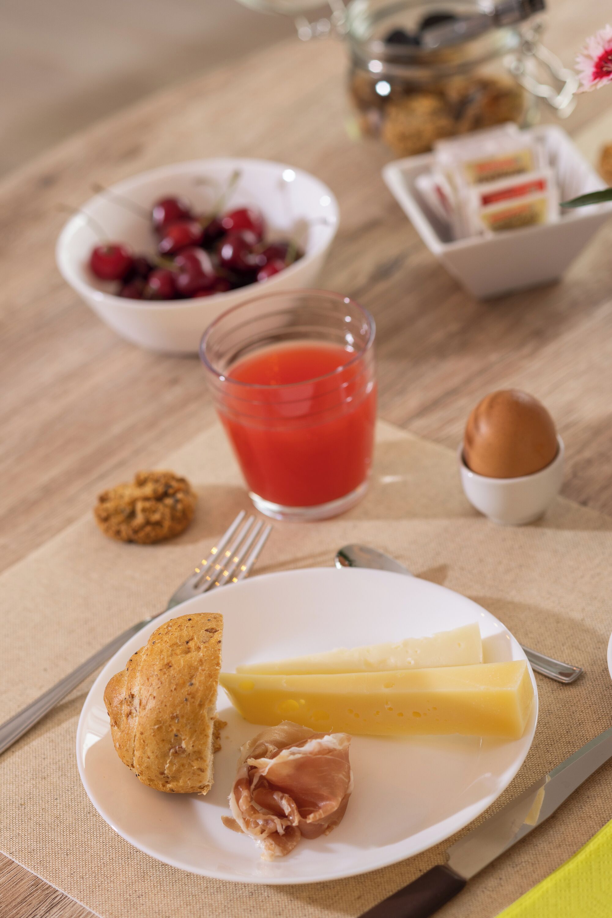 Uma mesa de café da manhã com um prato de comida, incluindo torradas, queijo e patê, um copo de suco vermelho e uma tigela de cerejas.