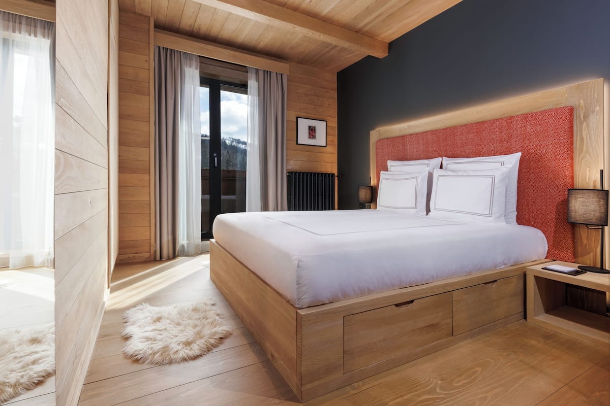 Uma cama de madeira com cabeceira vermelha, uma mesa de cabeceira com abajur e vista para o exterior por uma porta deslizante.