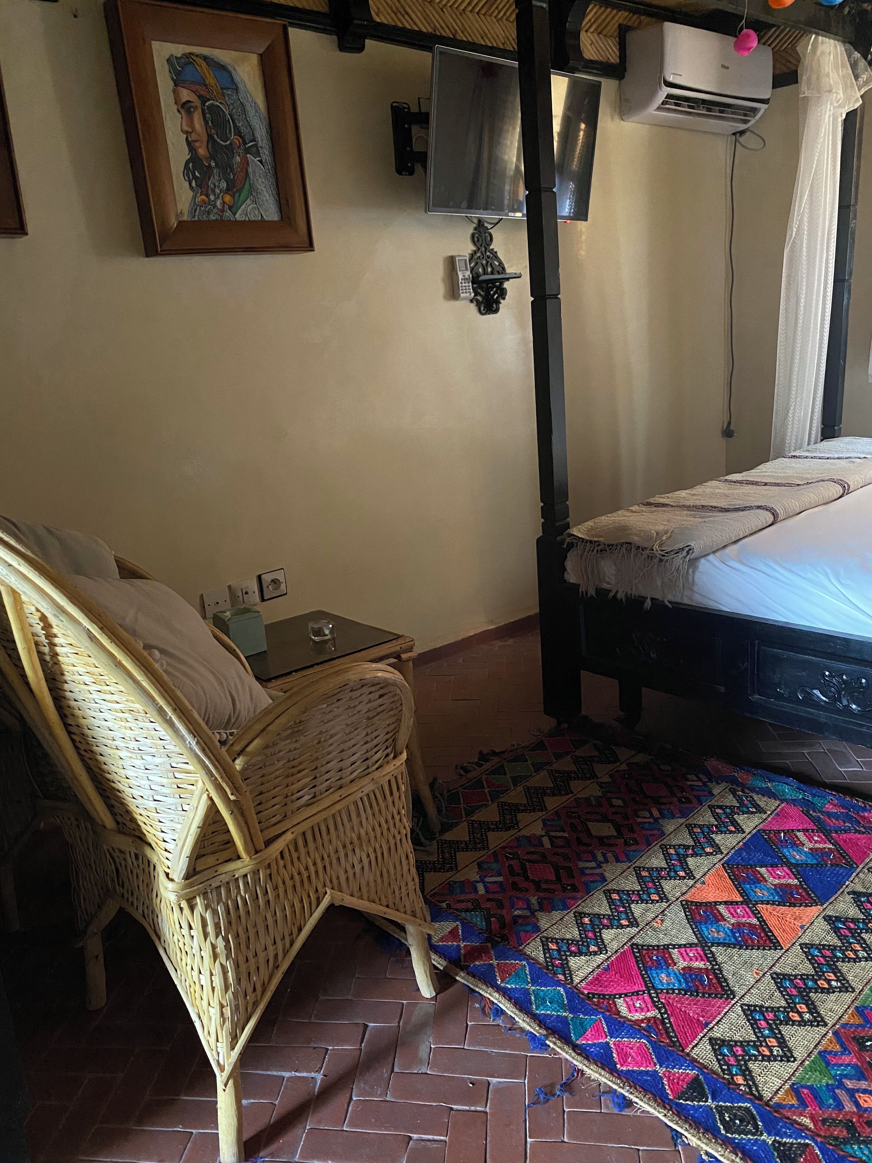 Quarto com cama, uma cadeira de vime, uma mesinha, um tapete colorido e um ar condicionado fixado na parede.
