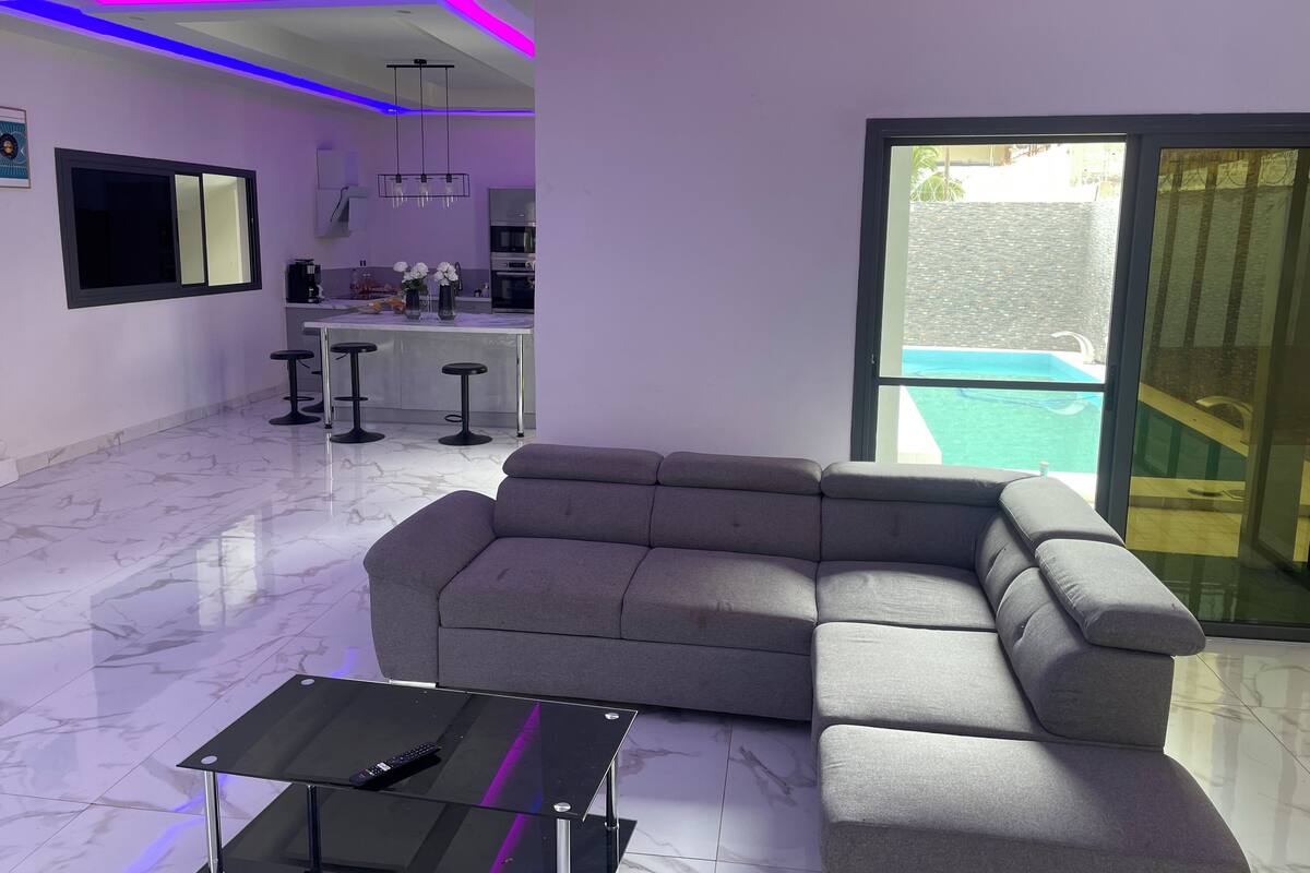 Uma sala de estar moderna com um sofá seccional cinza, uma mesa de centro de vidro e uma área de cozinha ao fundo.
