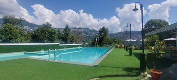 Piscina retangular com prancha de mergulho, rodeada por grama sintética, com vista para uma paisagem montanhosa e área de restaurante com mesas ao ar livre.