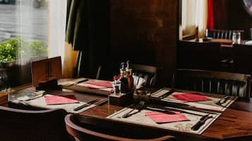 Interior de restaurante com parede de tijolos, janela com cortinas, mesa posta com guardanapos vermelhos e um violão no peitoril da janela.