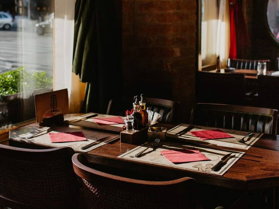 Interior de restaurante com parede de tijolos, janela com cortinas, mesa posta com guardanapos vermelhos e um violão no peitoril da janela.