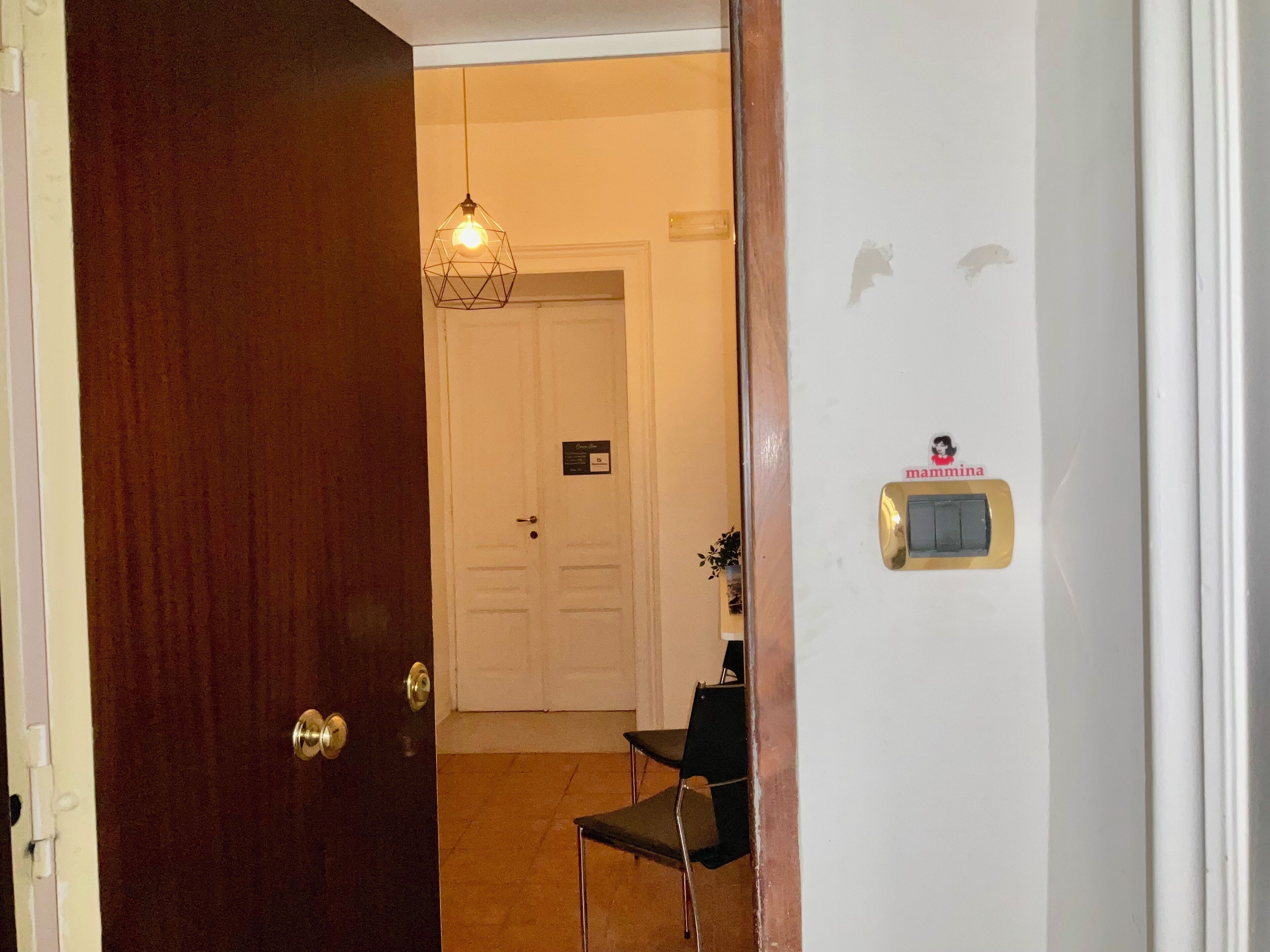 Um corredor com uma porta de madeira, uma porta branca, uma cadeira, um luminária fixada na parede e um termostato.