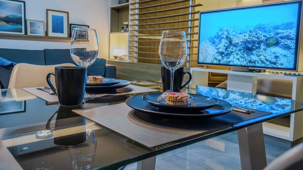 Uma sala de estar moderna com uma mesa de jantar de vidro, dois copos de vinho, um cupcake e uma TV de tela plana exibindo uma cena subaquática.