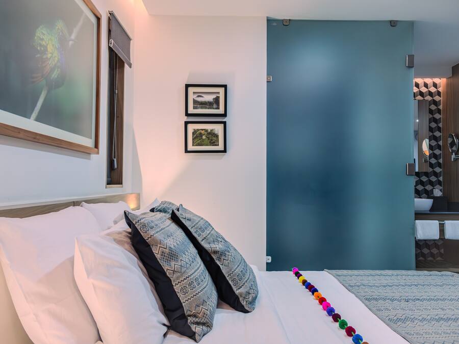 Uma cama bem arrumada com almofadas, um brinquedo pompom colorido e duas fotos emolduradas na parede.
