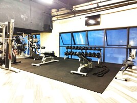 Uma academia com equipamentos de ginástica, suportes para pesos e uma grande janela com cortinas azuis.