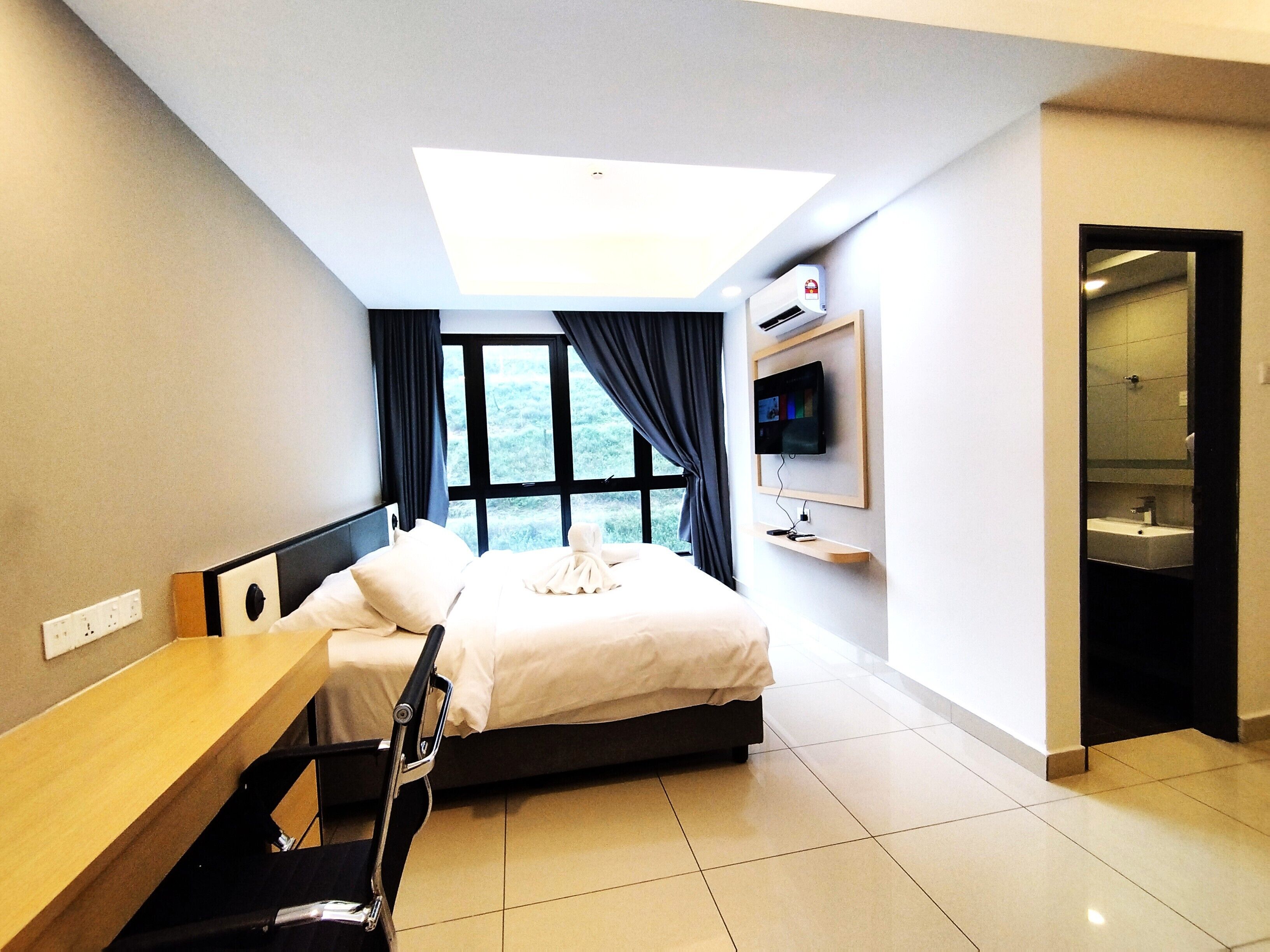 Quarto de hotel com cama, escrivaninha, televisão na parede e janela com cortinas.