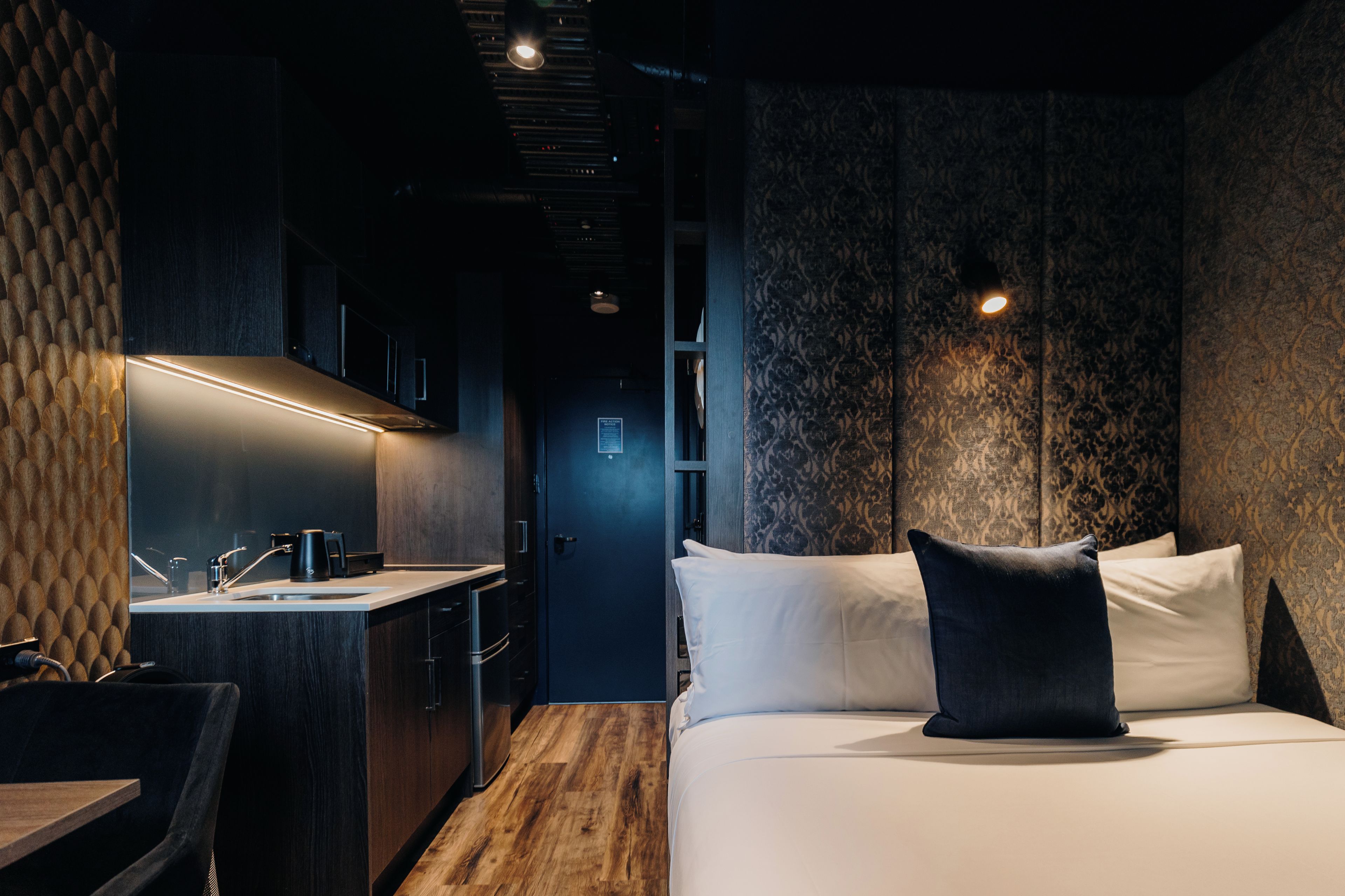 Quarto de hotel moderno com piso de madeira, cama com roupa de cama branca, parede com padrão escuro e uma pequena cozinha com pia e cafeteira.