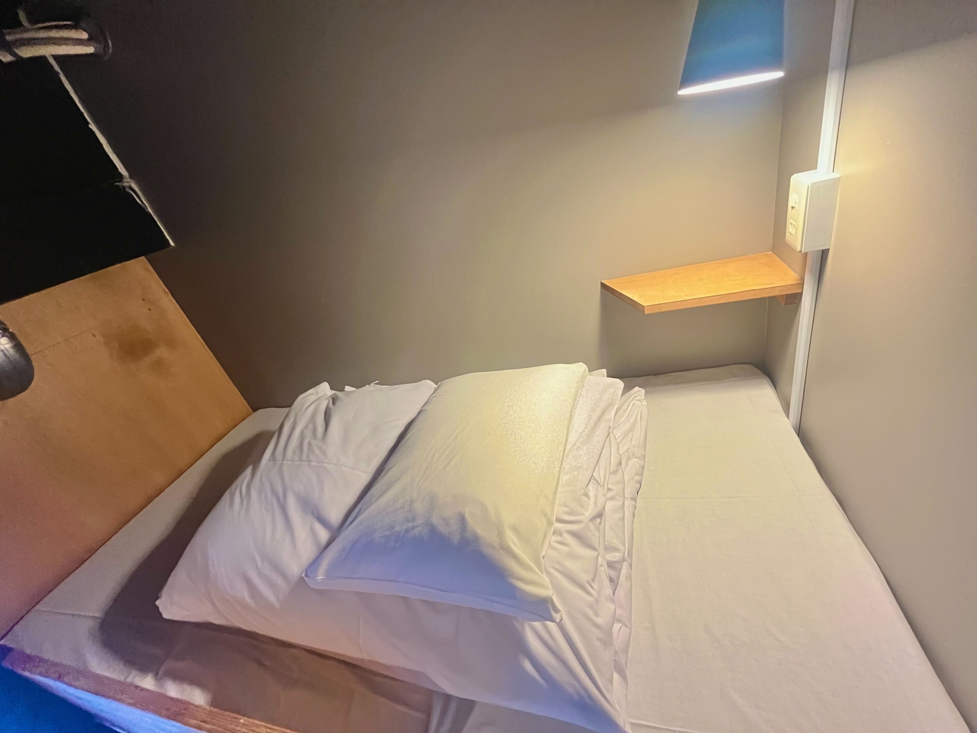 Uma cama com roupa de cama e almofadas brancas, cabeceira de madeira, mesa de cabeceira com abajur e prateleira fixada na parede.