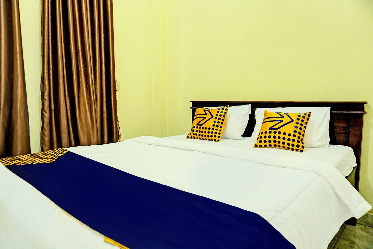 Cama com roupa de cama branca, duas almofadas com padrões amarelos e pretos, e um cobertor azul.
