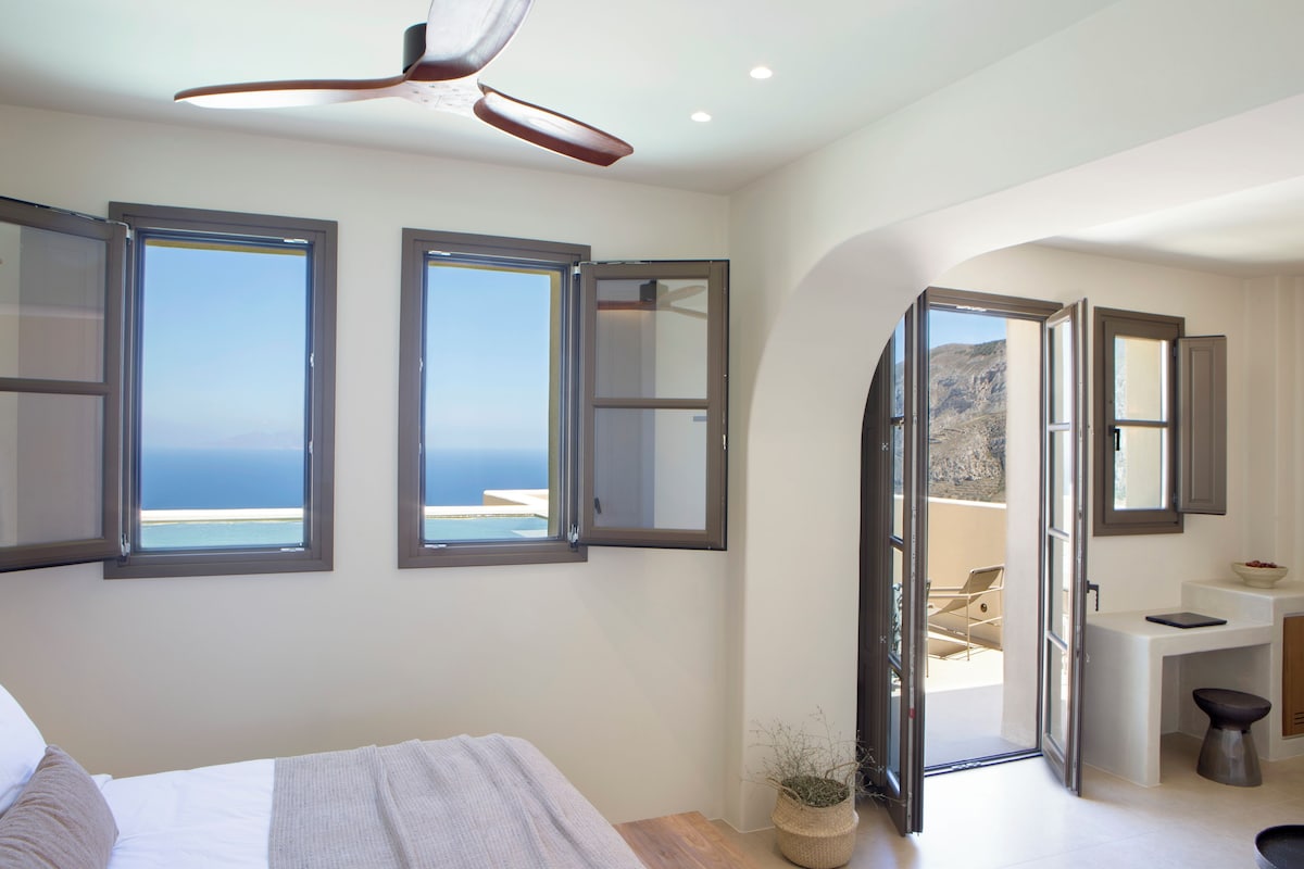 Quarto com cama, ventilador de teto e vista para o mar através de janelas abertas.
