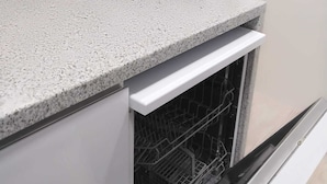 Uma cozinha com bancada de granito, máquina de lavar louça e um sofá cinza ao fundo.