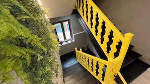 Uma escadaria com corrimão amarelo, um vaso de planta no patamar e uma janela com flores.