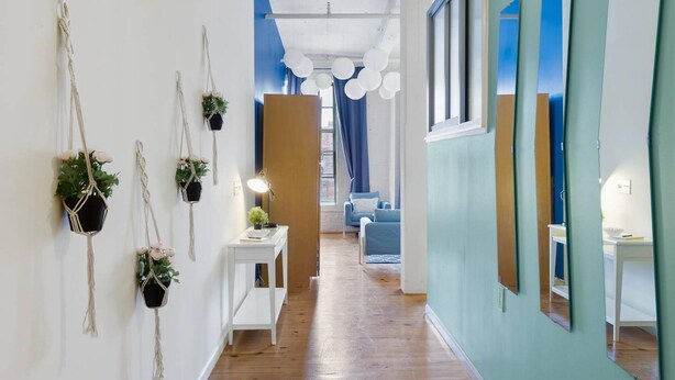 Um corredor com piso de madeira, paredes brancas e uma parede azul de destaque. Há suportes de plantas em macramé na parede esquerda e uma mesa branca com abajur no centro. Um sofá azul e uma cadeira branca são visíveis ao fundo.