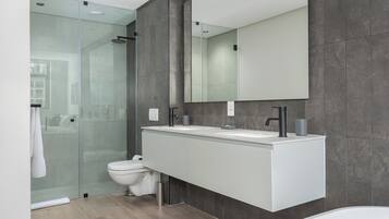 Uma casa de banho moderna com cabine de duche em vidro, sanita branca, espelho grande e toucador branco com lavatório e duas torneiras.