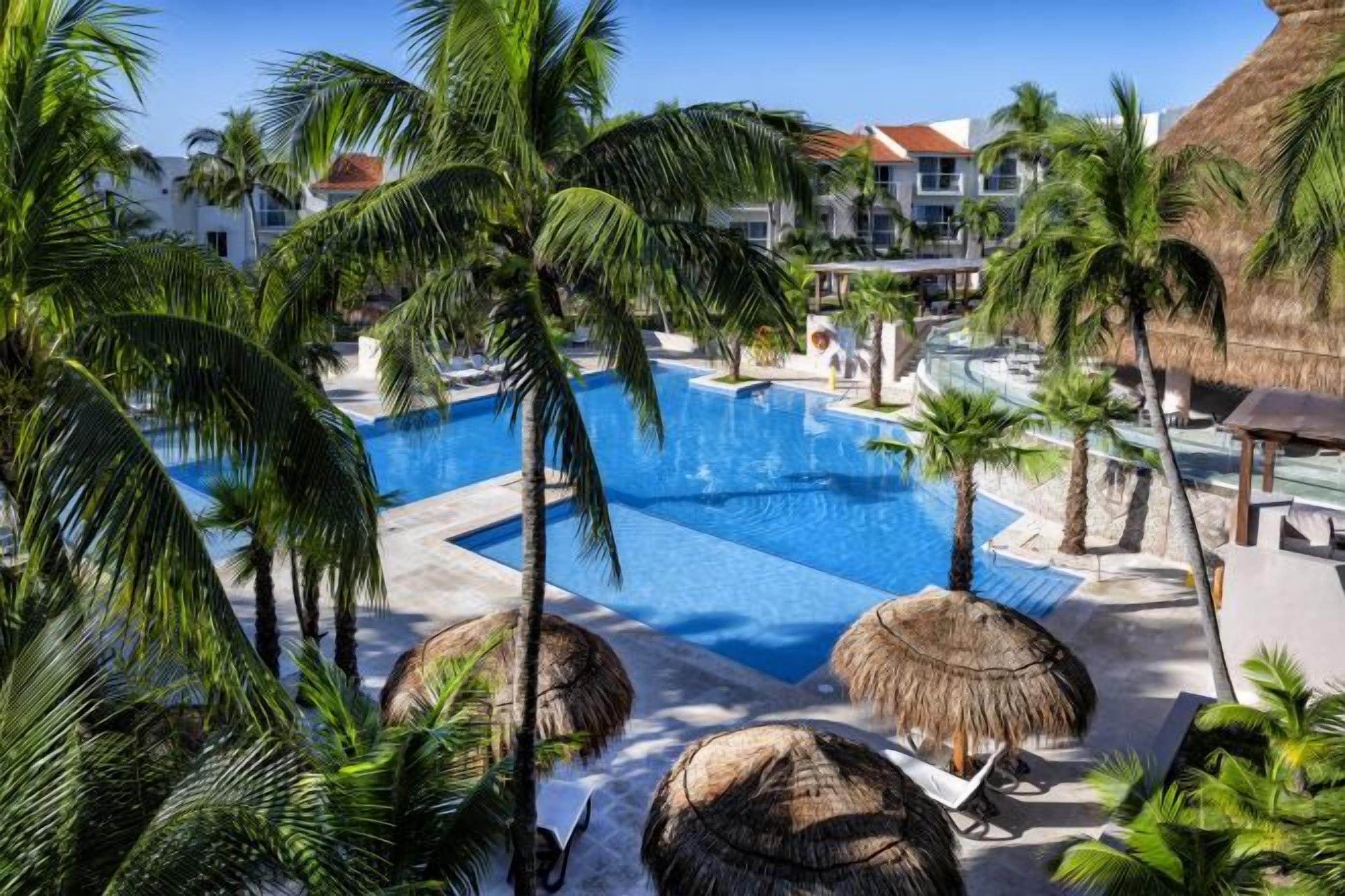 Área de piscina em resort com palmeiras, guarda-sóis de palha e uma piscina azul cristalina.