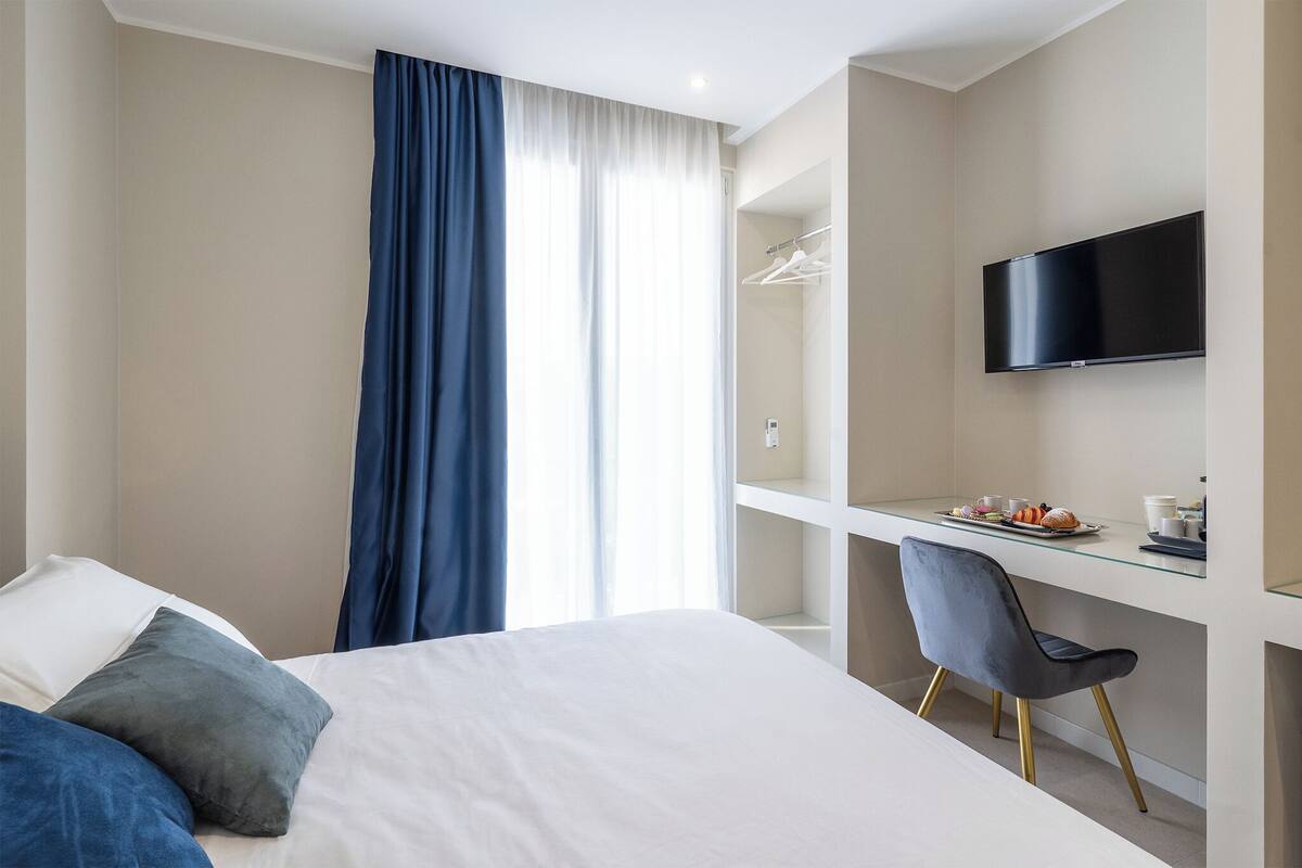 Quarto de hotel moderno com cama, secretária com cadeira, televisão e janela com cortinas.
