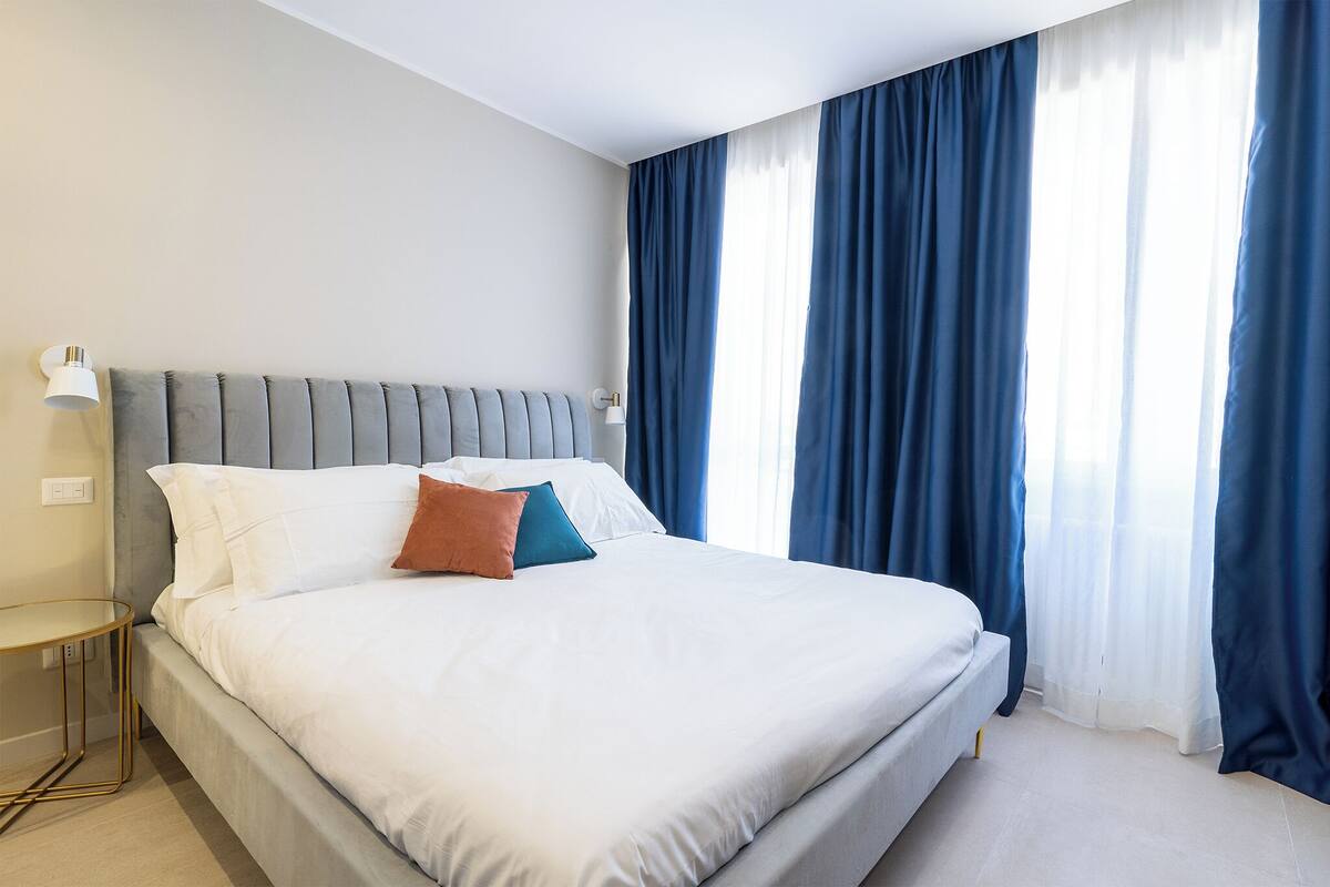 Cama bem arrumada com travesseiros brancos e azuis, cabeceira cinza e mesa de cabeceira com abajur.
