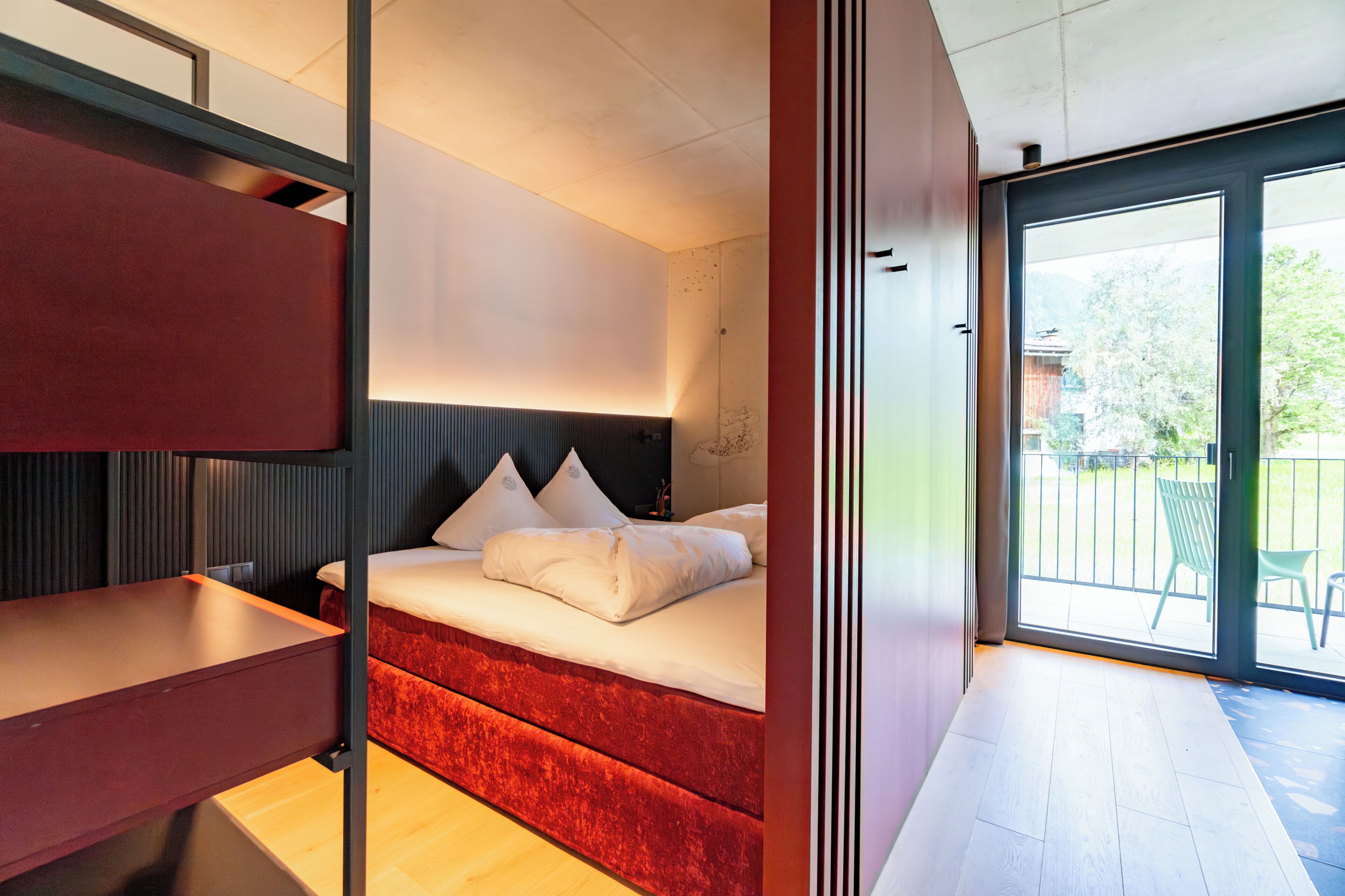 Quarto moderno com cama vermelha, escrivaninha de madeira e porta de vidro deslizante para uma varanda.