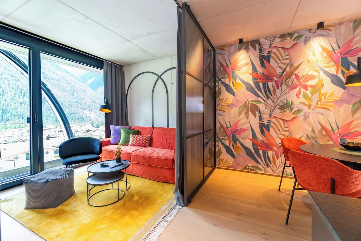 Uma sala de estar moderna com uma janela grande, um sofá vermelho, uma cadeira preta, uma mesa redonda pequena e um papel de parede tropical vibrante.