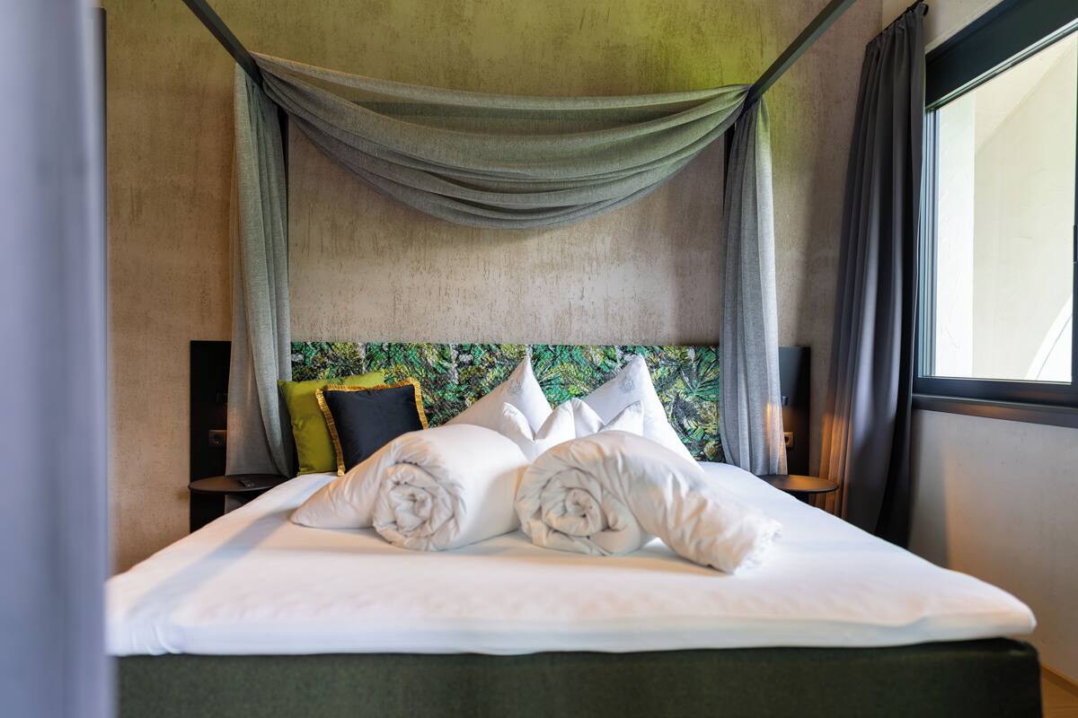 Cama bem arrumada com toalhas dobradas, um travesseiro verde e cabeceira com padrão de folhas.