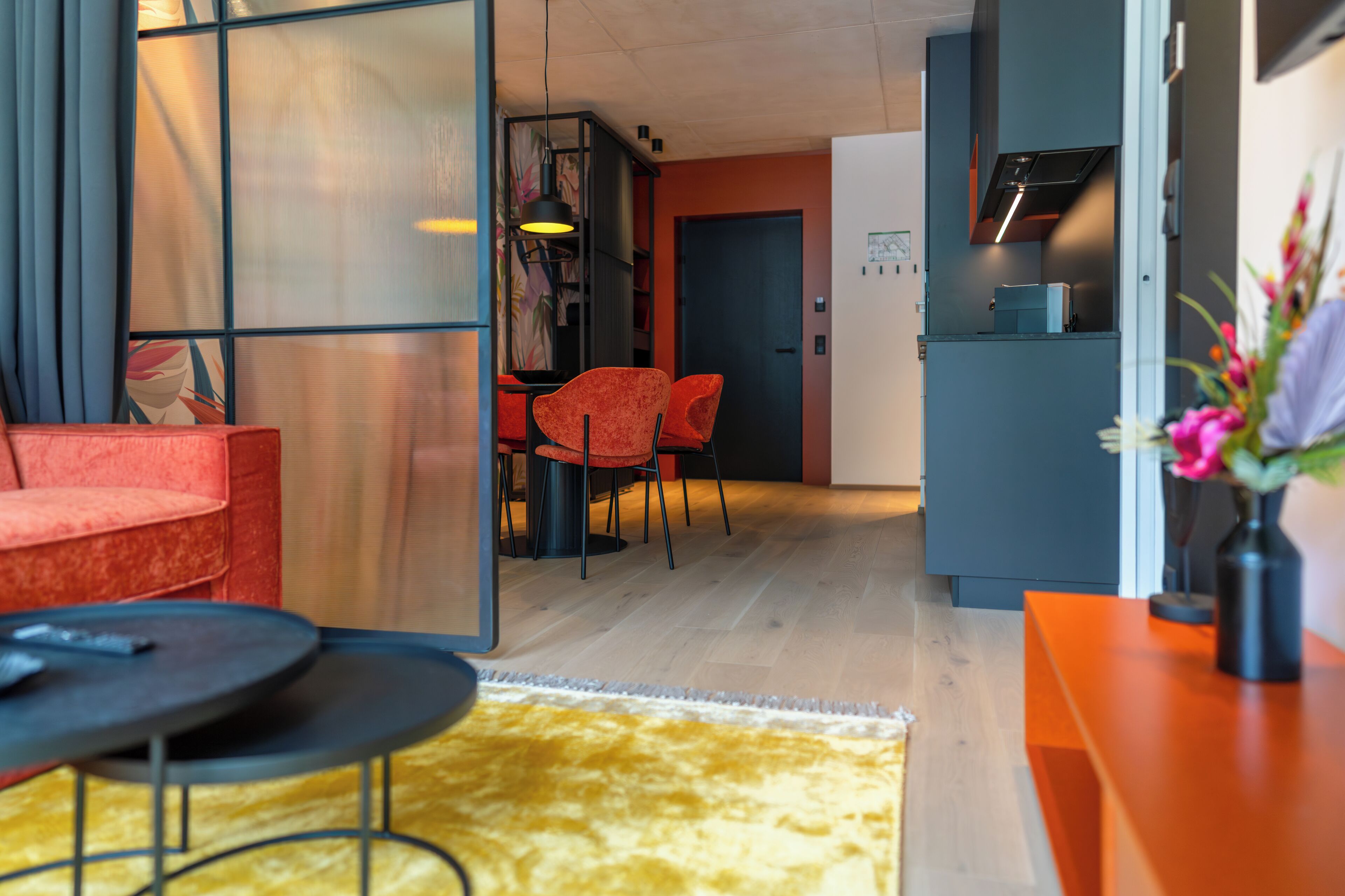 Uma sala de estar moderna com um sofá vermelho, uma mesa de centro preta e um tapete amarelo. Há uma área de jantar com cadeiras vermelhas e uma cozinha com geladeira e fogão.