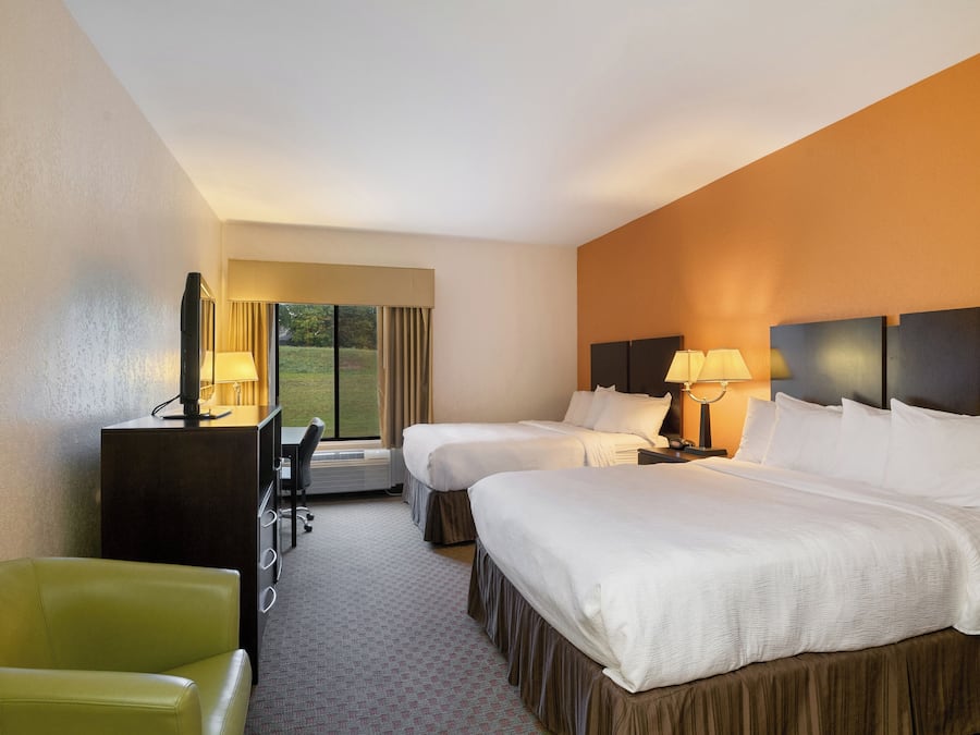 Quarto de hotel com duas camas, uma mesa, uma cadeira e uma janela grande com cortinas.
