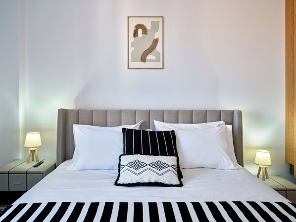 Cama bem arrumada com colcha listrada preta e branca, cabeceira estofada cinza e duas luminárias de mesa em mesas de cabeceira de madeira.