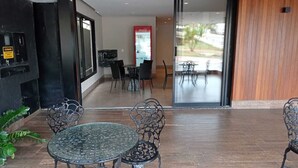 Uma área de estar interna moderna com uma mesa de vidro e cadeiras de metal, um refrigerador Coca-Cola e vista para o pátio externo.