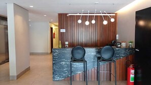 Uma área de recepção moderna com balcão de mármore, dois banquetas pretas, painel de parede de madeira e teto com luminárias pendentes.