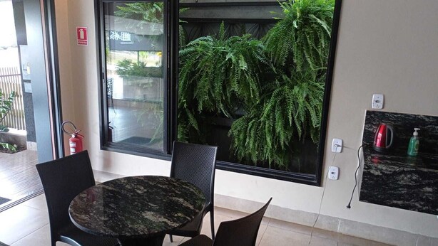 Um quarto com uma mesa redonda e cadeiras, uma janela grande com vista para samambaias, um extintor de incêndio e uma tomada elétrica na parede.