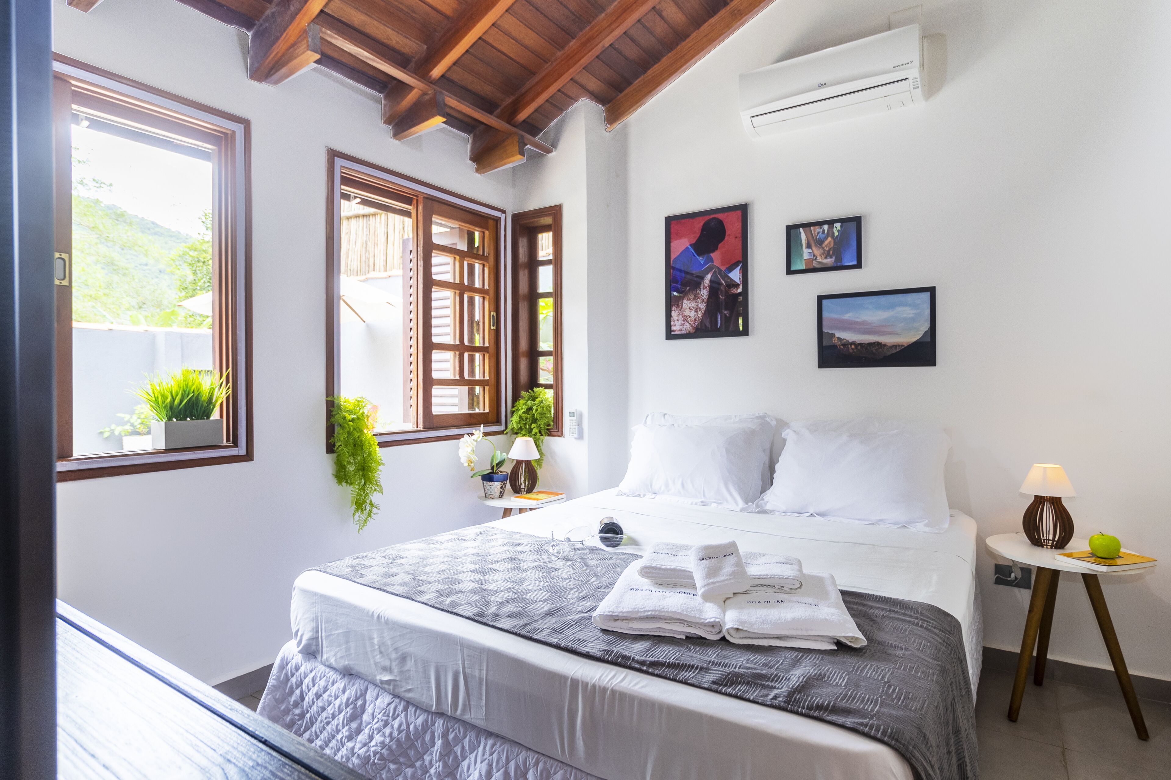 Quarto com cama, duas janelas, ar-condicionado na parede e uma mesinha de cabeceira com abajur e uma maçã.