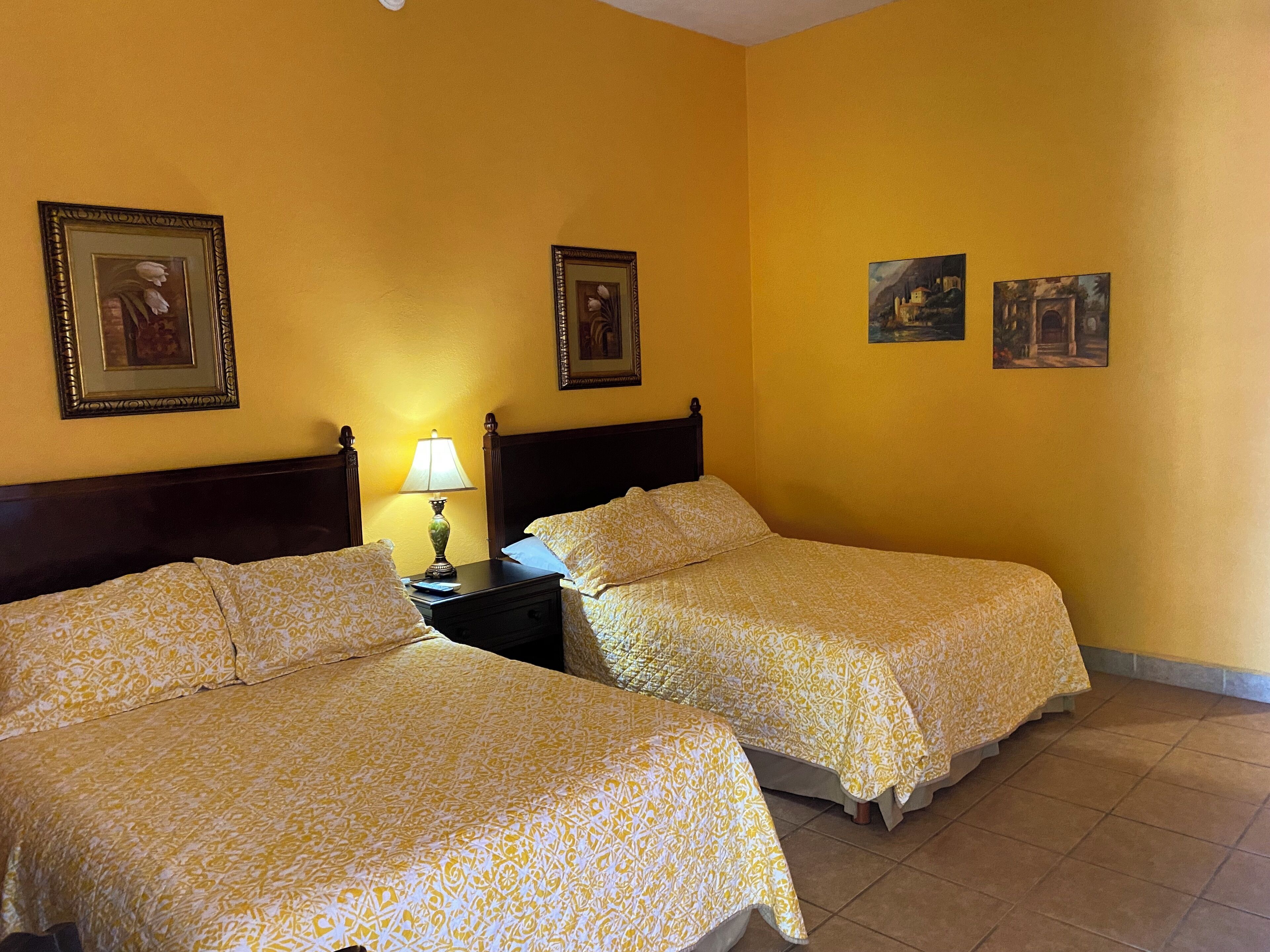 Quarto de hotel com duas camas, uma mesa de cabeceira com abajur e três quadros na parede.