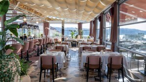 Um restaurante com mesas postas para refeições, grandes janelas com vista e um teto com design ondulado.